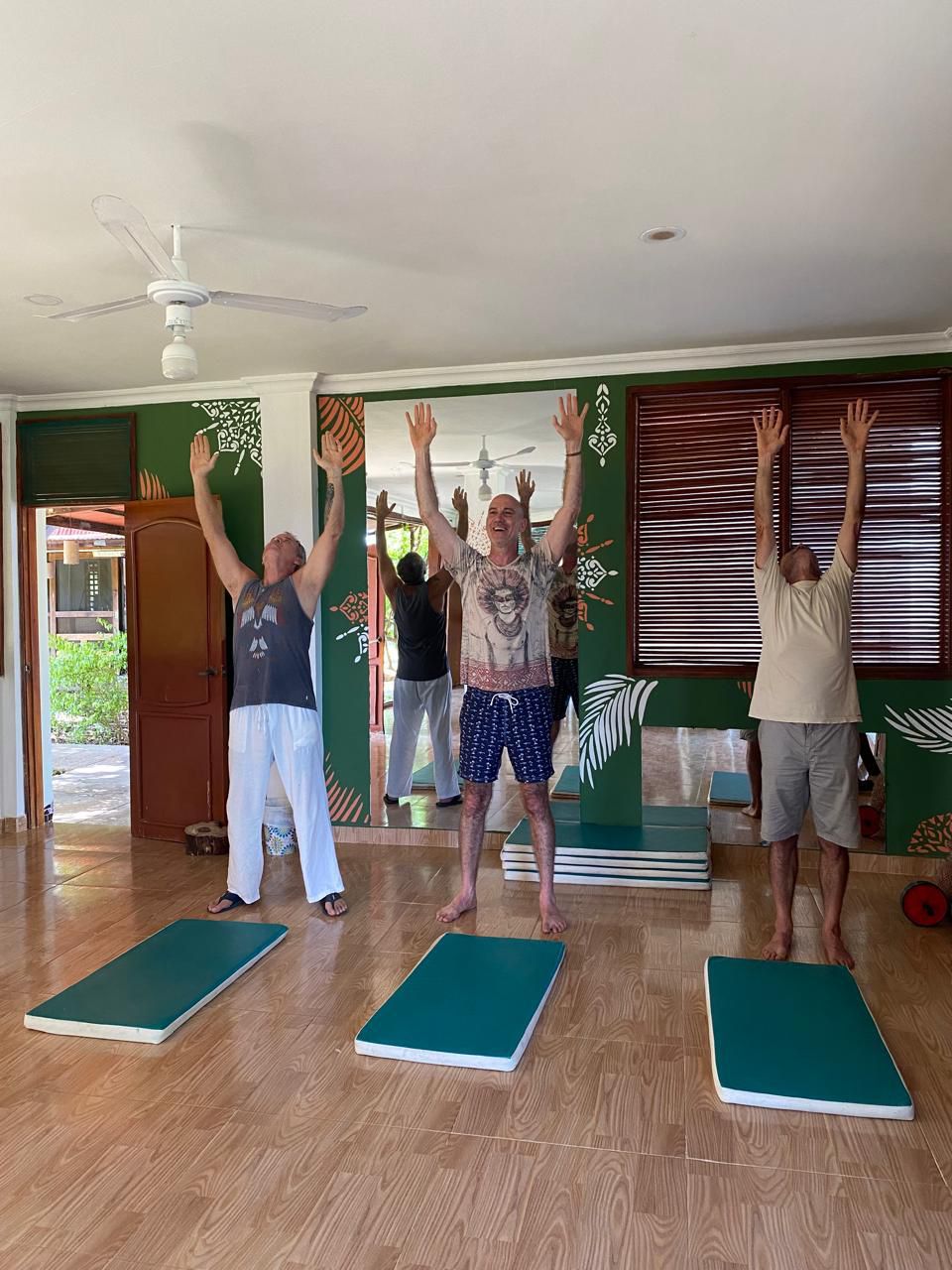Aula de yoga em um espaço com piso de madeira, tapetes verdes e uma janela grande com persianas.