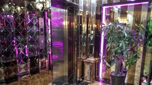Um saguão de hotel com um elevador de vidro, uma mesa de recepção e um vaso de planta.