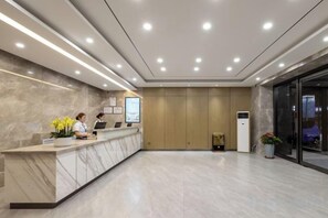 Uma área de recepção de hotel com balcão de mármore, tela digital e mesa de atendimento.