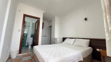 Quarto com cama, mesa de cabeceira, luminária na parede e banheiro visível através de uma porta aberta.