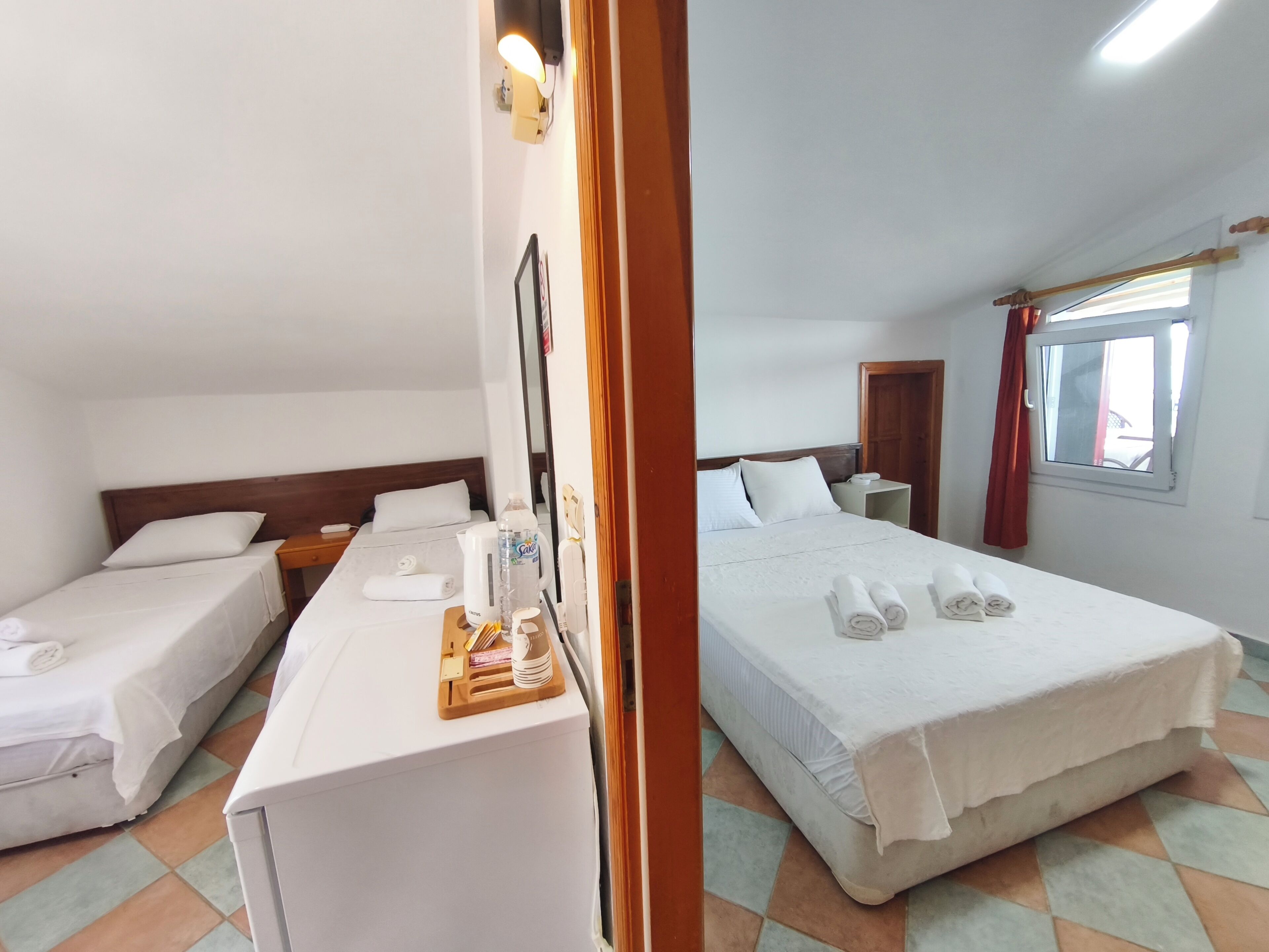 Quarto de hotel com duas camas, uma mesinha com chaleira, espelho e janela com cortinas.