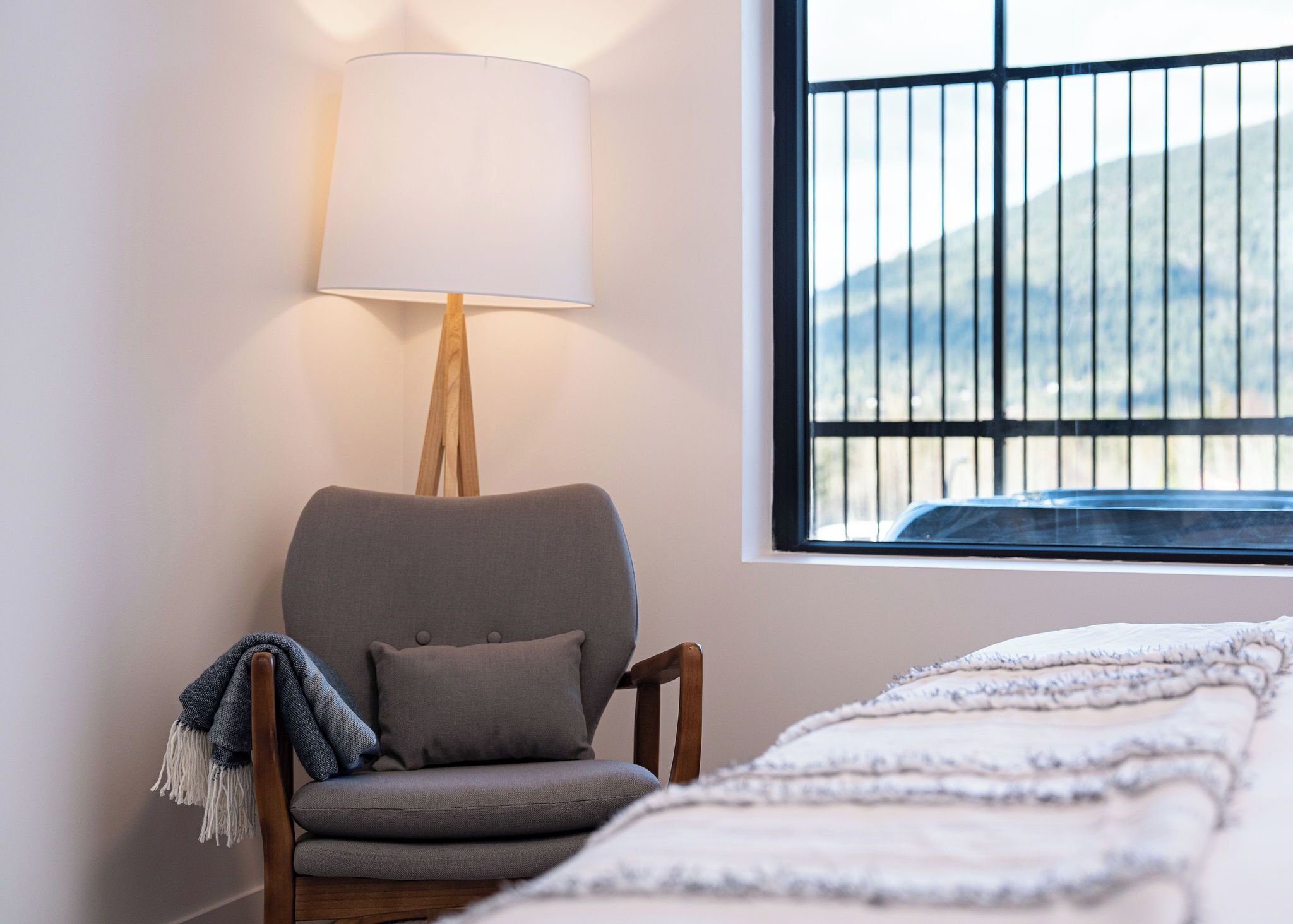Quarto com poltrona cinzenta, luminária de chão branca, janela com vista para montanhas e cama com manta branca.