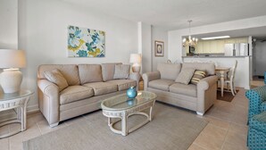 Uma sala de estar com dois sofás bege, uma mesa de centro de vidro e um espaço de jantar com mesa e cadeiras.