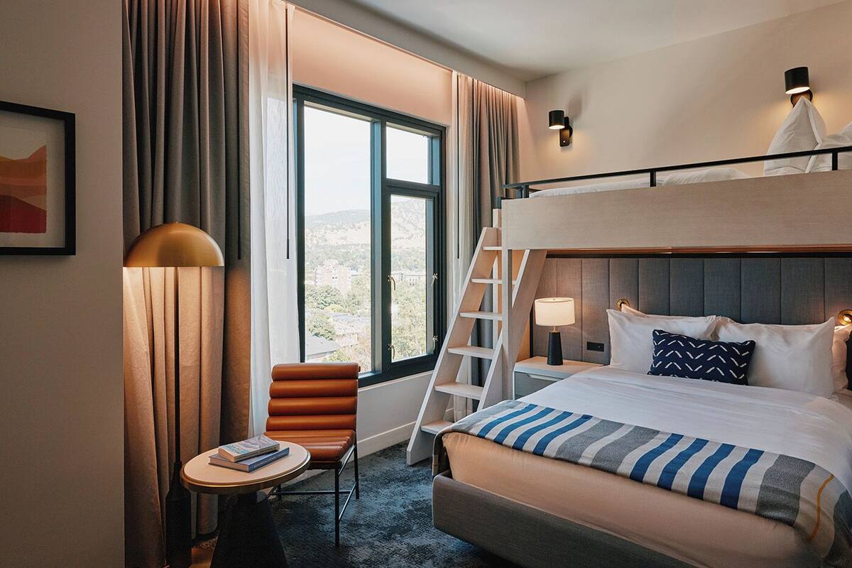 Quarto de hotel com beliche, escrivaninha com cadeira, janela com cortinas e luminária.