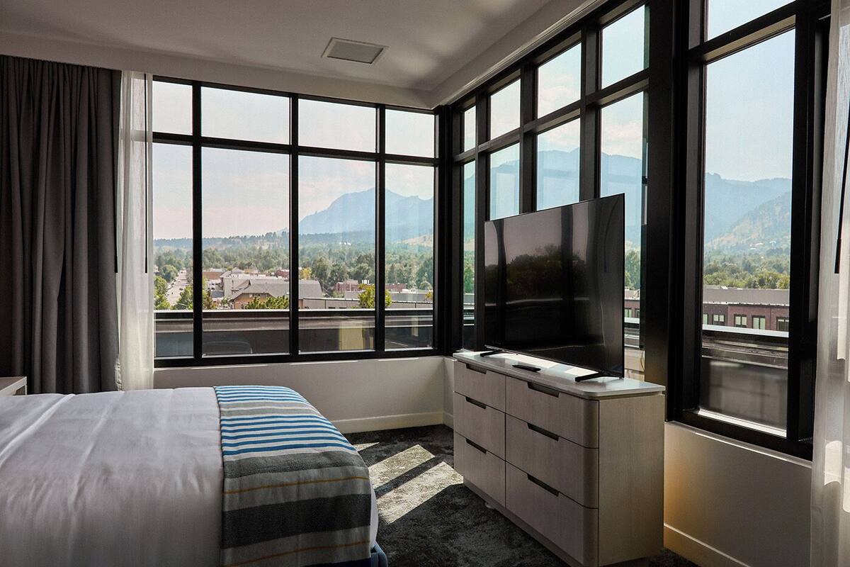 Quarto de hotel com cama, televisão, penteadeira e grandes janelas com vista para as montanhas.