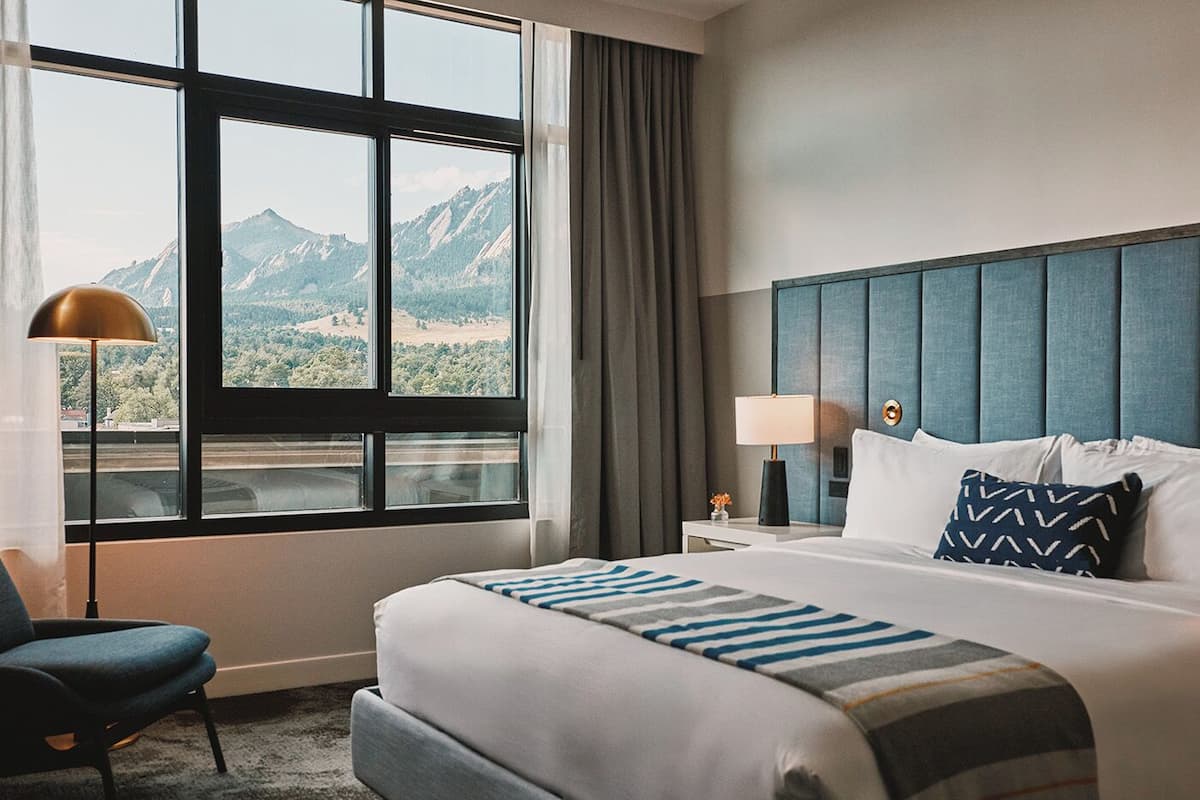 Quarto de hotel com uma grande janela que oferece vista para as montanhas, uma cama com manta listrada, uma cadeira azul e uma mesa de cabeceira com abajur.