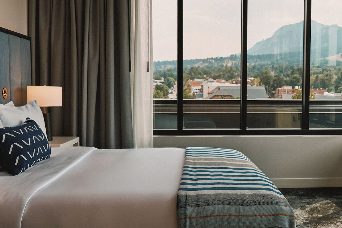 Quarto de hotel com uma grande janela, cama com roupa de cama listrada, luminária e vista para uma paisagem urbana e montanhas.