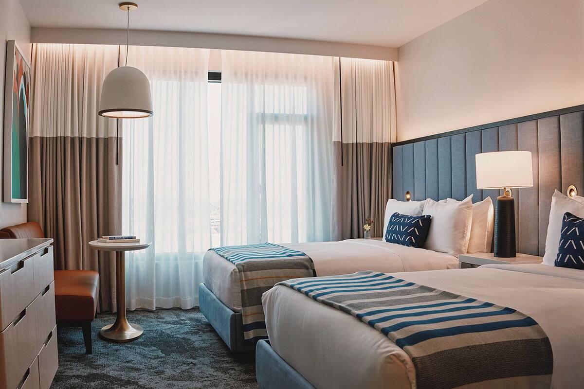 Quarto de hotel com duas camas, uma escrivaninha, uma cadeira, um abajur e vista para o exterior através de cortinas finas.