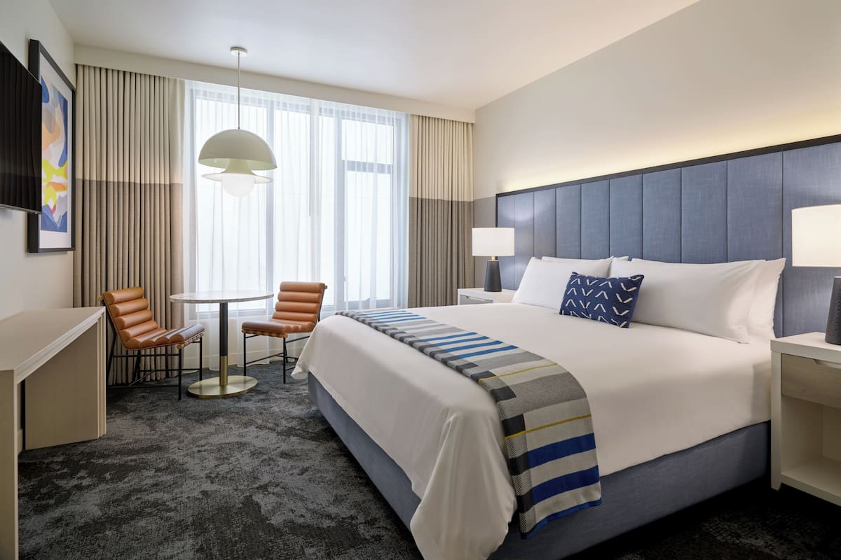 Quarto de hotel com uma cama grande, uma escrivaninha, uma cadeira, uma luminária e uma TV na parede.