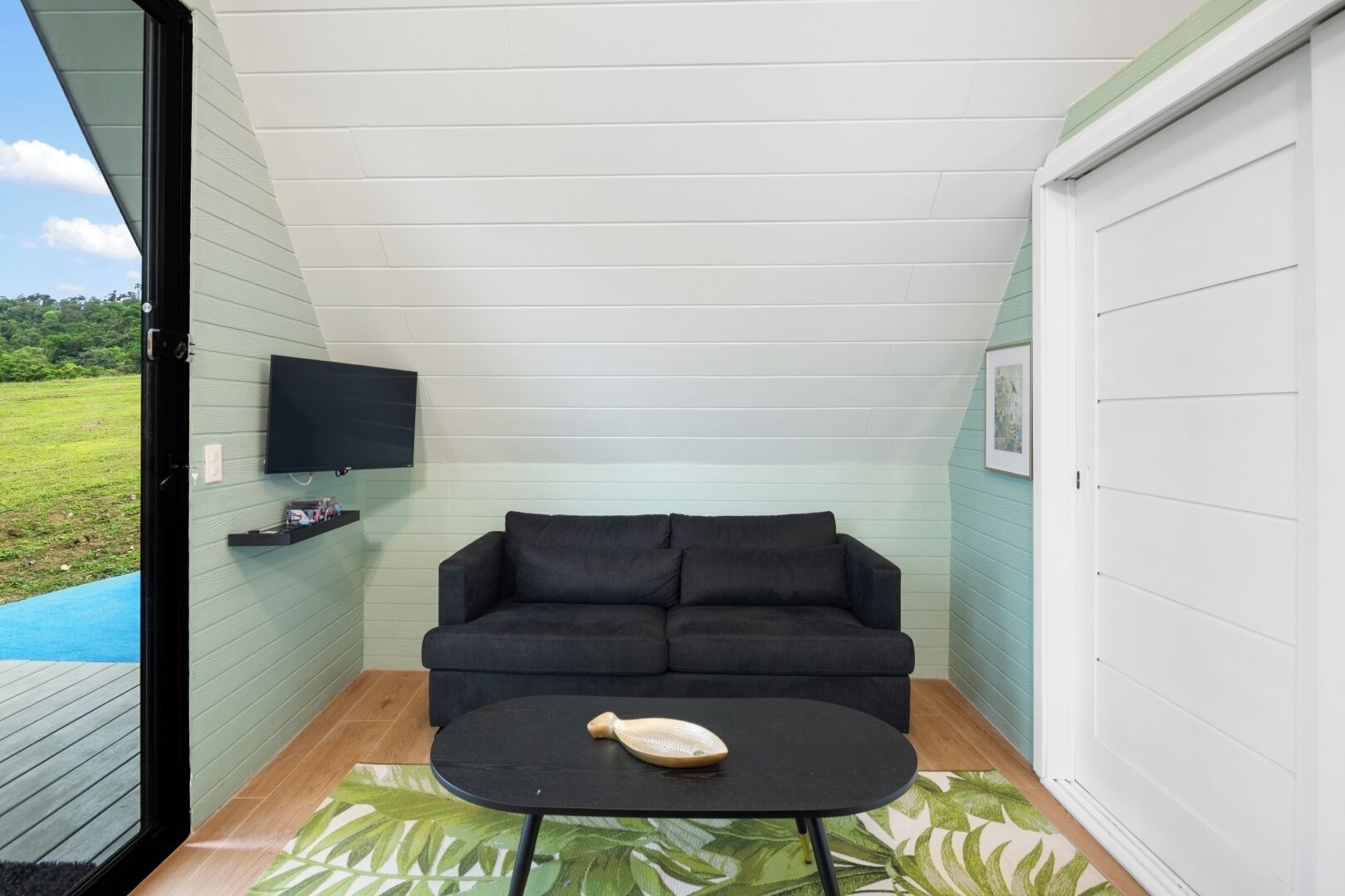 Quarto aconchegante com sofá preto, mesa redonda, TV na parede e vista para um campo verde através de uma porta de correr.