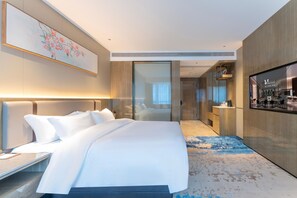 Quarto de hotel moderno com uma cama grande, uma mesa de escritório, uma televisão de tela plana e um banheiro visível através de uma porta de vidro.