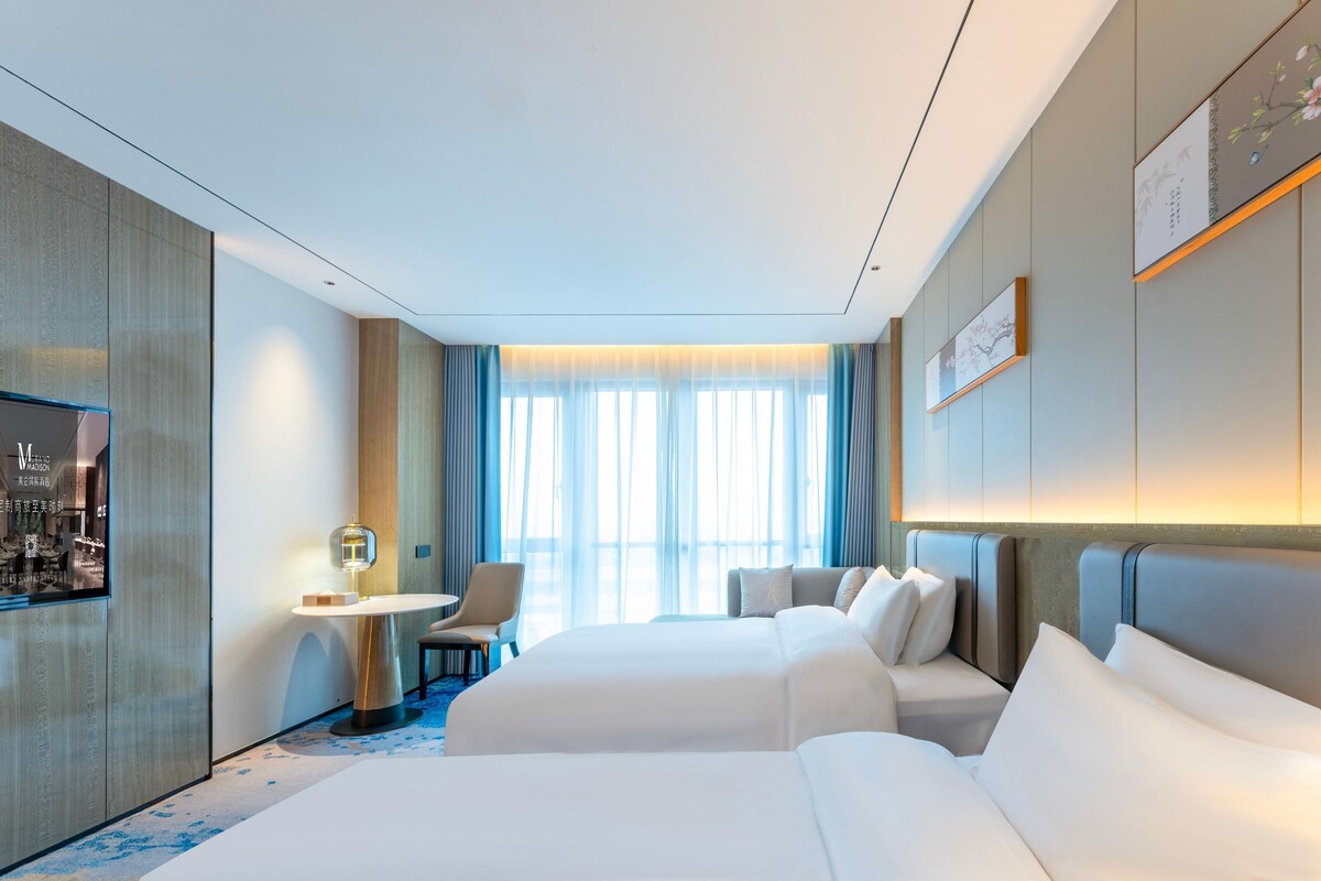 Quarto de hotel com duas camas, uma escrivaninha, uma cadeira e uma janela grande com cortinas.