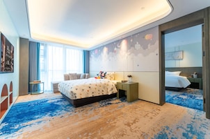 Quarto de hotel moderno com uma cama grande, uma mesinha de cabeceira, uma pequena mesa e uma janela ampla.