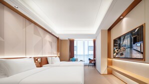 Quarto de hotel com duas camas, área de estar com mesa e cadeiras, uma janela grande com cortinas e uma TV fixada na parede.