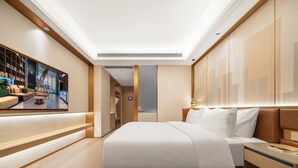 Quarto de hotel moderno com uma cama grande, uma mesinha de cabeceira, um criado-mudo, um espaço de estar com sofá e mesa de centro, e uma janela ampla com vista para a cidade.