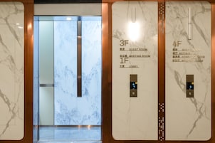 Interior de elevador com paredes e chão de mármore, visor digital exibindo números de andares e etiquetas para quartos de hóspedes e salas de reunião.