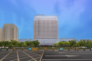 Um hotel moderno com fachada de vidro, vários andares e estacionamento em frente.