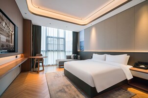 Quarto de hotel moderno com uma cama grande, área de estar, televisão e vista para o mar pelas janelas.