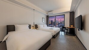 Quarto de hotel com duas camas, televisão, escrivaninha com cadeira e uma janela grande com vista para a cidade.