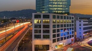 Um edifício moderno de vários andares com fachada de vidro e janelas iluminadas, localizado ao lado de uma rua movimentada com rastros de luz indicando o tráfego.
