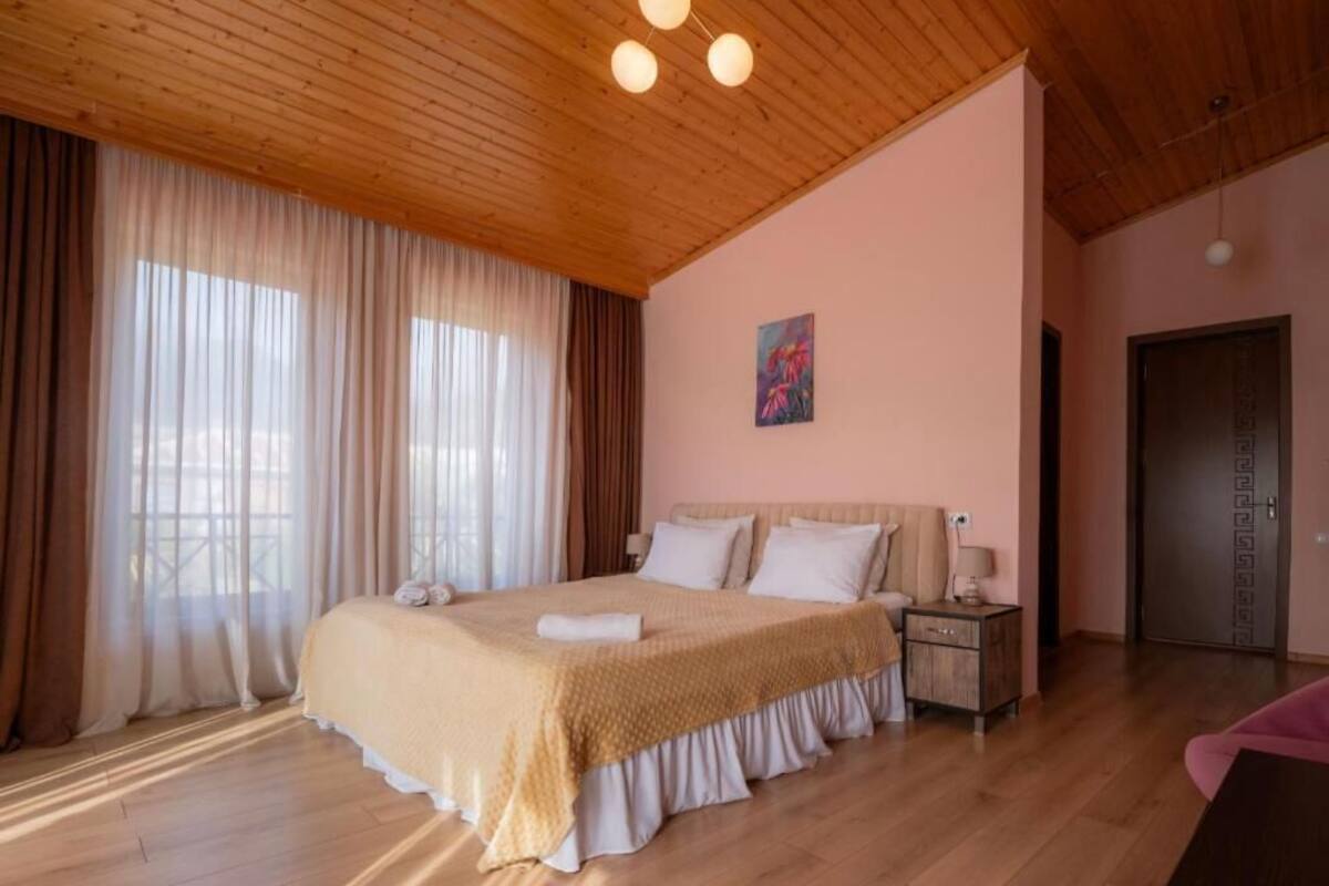 Quarto com teto de madeira, cama com colcha bege, mesa de cabeceira, um quadro na parede e uma porta.