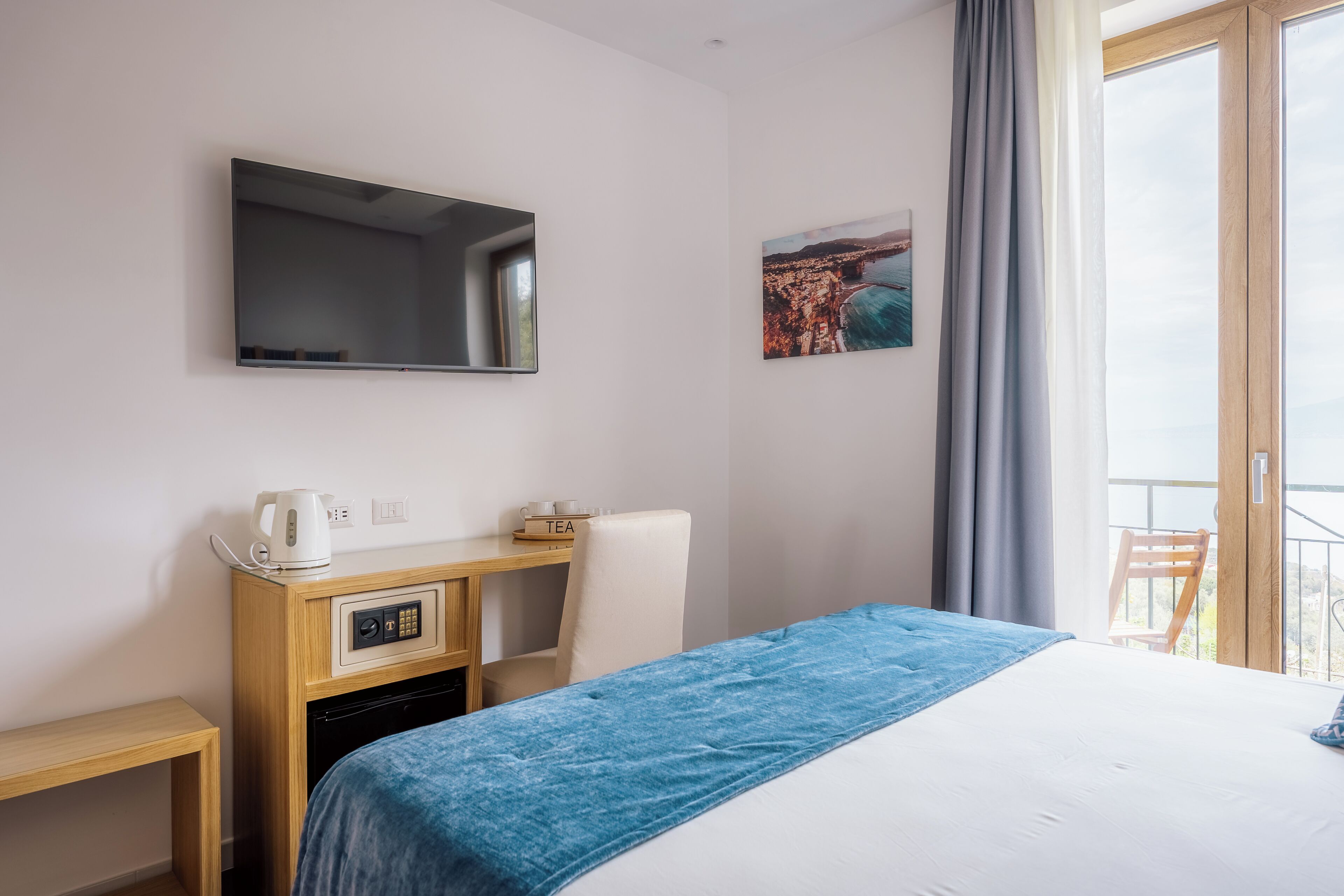 Quarto de hotel com uma cama, uma escrivaninha com chaleira, uma cadeira, televisão e varanda com vista.