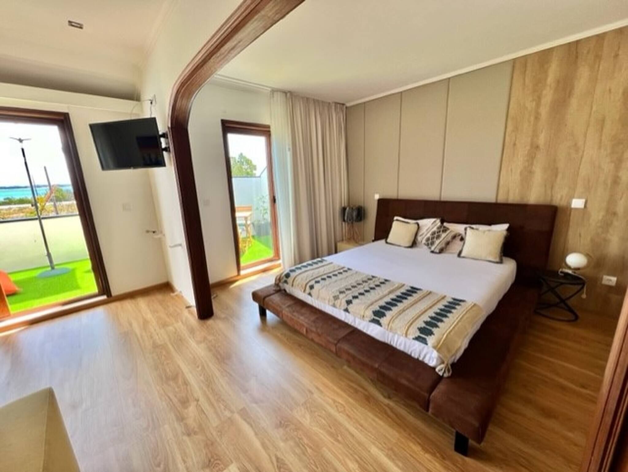 Um quarto espaçoso com uma cama grande, piso de madeira, televisão de tela plana na parede e uma porta de vidro deslizante que dá para uma varanda com vista para o mar.