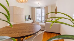 Uma sala de estar moderna com uma mesa de madeira redonda, sofá branco e uma televisão sobre um móvel.
