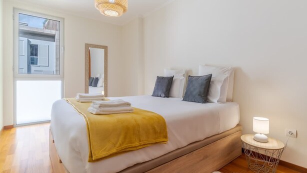 Uma cama bem arrumada com um cobertor amarelo, uma luminária de cabeceira e vista para o exterior através de uma janela.