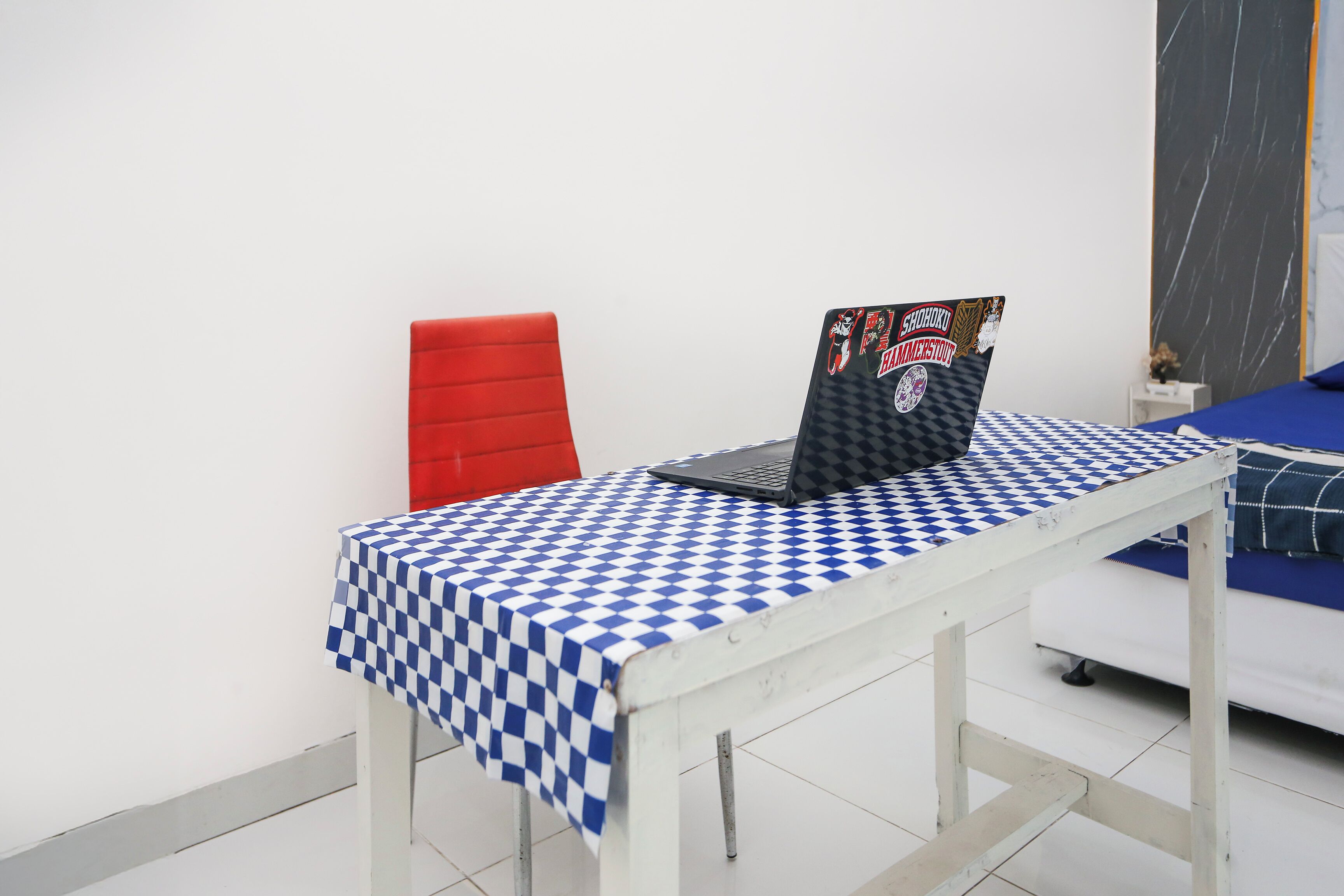 Um quarto com uma mesa xadrez, um laptop, uma cadeira vermelha e uma cama com cobertura azul.