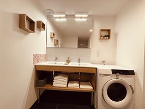 Uma casa de banho moderna com uma sanita dupla, espelho, máquina de lavar roupa embutida e uma prateleira com toalhas dobradas.