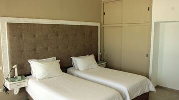 Quarto de hotel com duas camas de solteiro, uma cabeceira grande e acolchoada, uma mesa de cabeceira com luminária e um guarda-roupa.