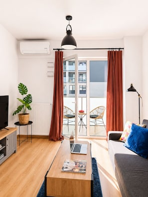 Uma sala de estar moderna com um sofá, uma mesa de centro com um laptop, uma TV, uma planta em vaso e vista para uma varanda com cadeiras.