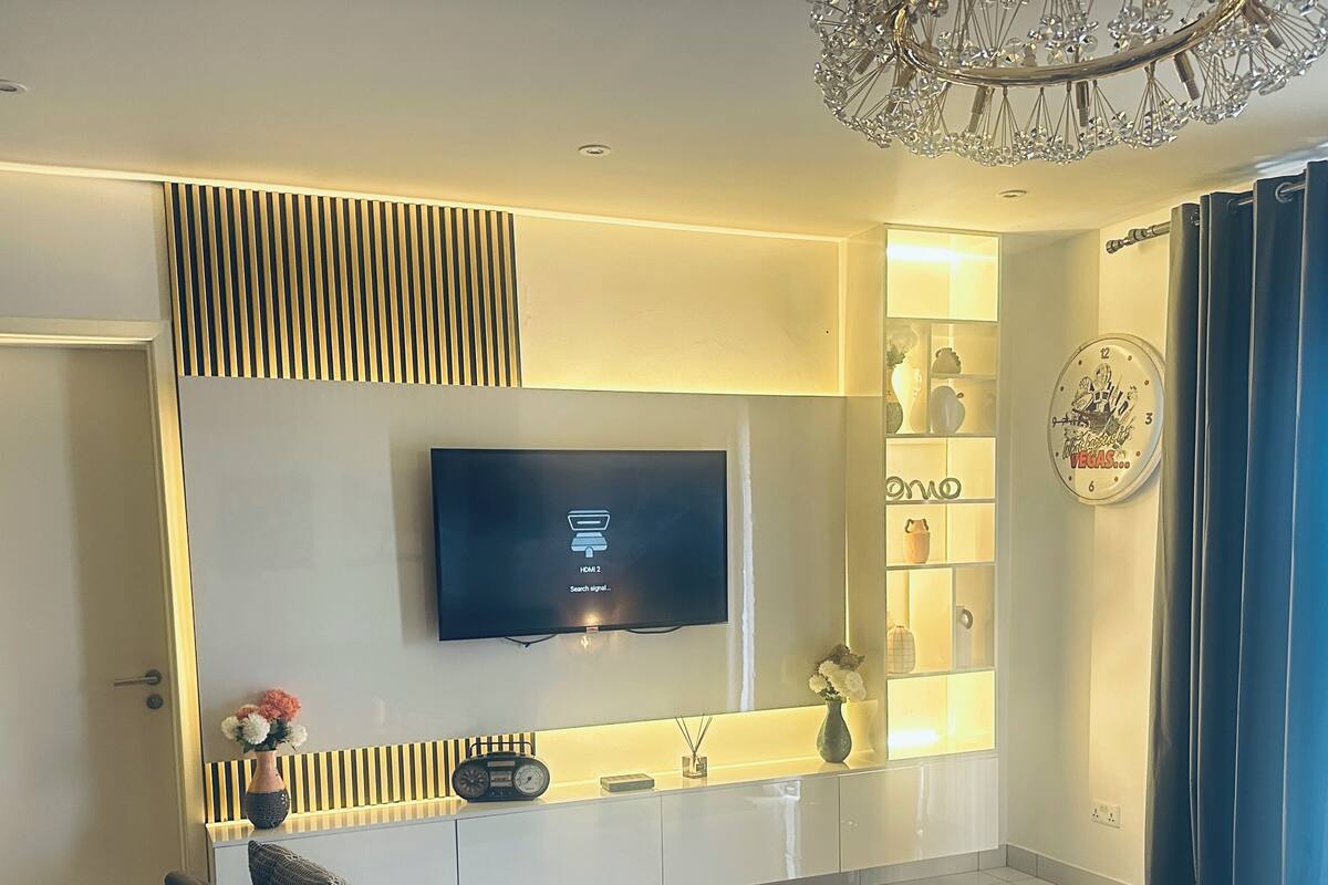 Uma sala de estar moderna com uma televisão de tela plana na parede, um sofá preto, uma mesa de centro e um relógio decorativo.