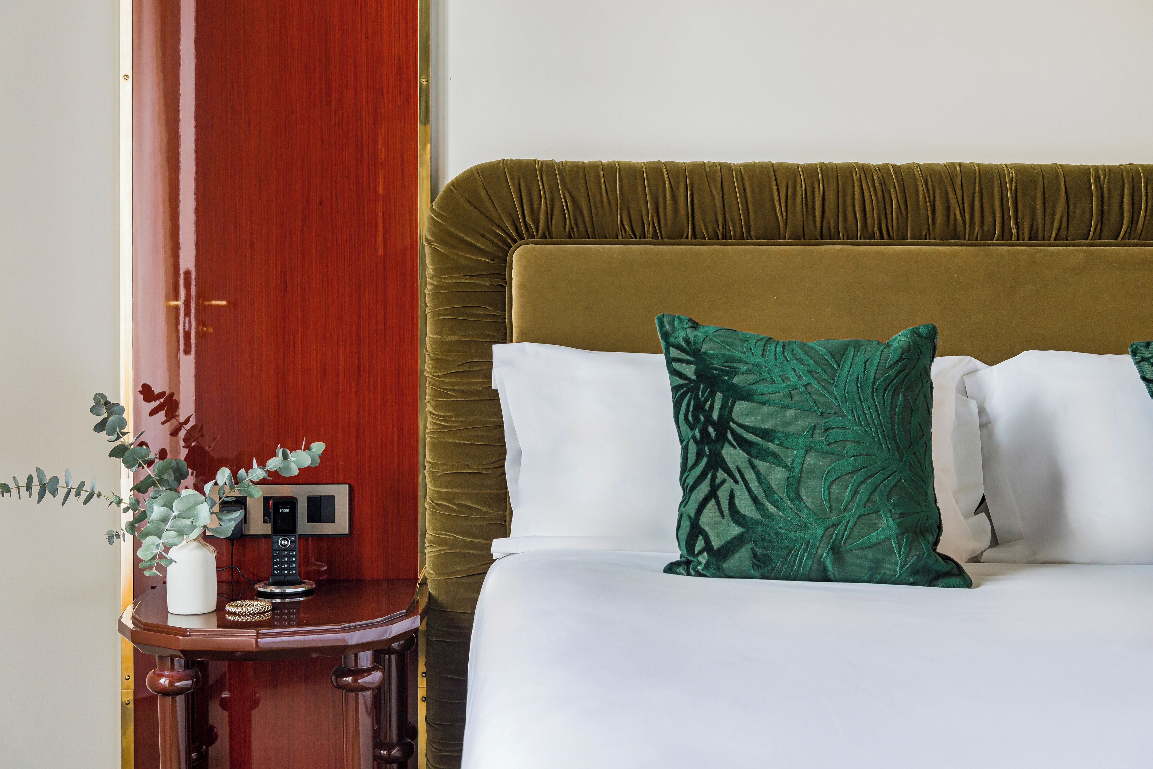 Uma cama bem arrumada com um travesseiro verde e um travesseiro branco, uma mesa de cabeceira de madeira com um vaso e um controle remoto, e uma porta de madeira com maçaneta.
