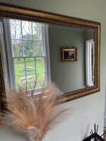 Um espelho com moldura dourada reflete uma janela com vista para uma área gramada e uma mesa de piquenique. Uma pequena moldura com um quadro está pendurada na parede.