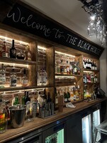 Uma área de bar com um letreiro 'Welcome to The Black Horse', prateleiras com diversas garrafas de bebidas e um balcão com copos e um caixa registradora.