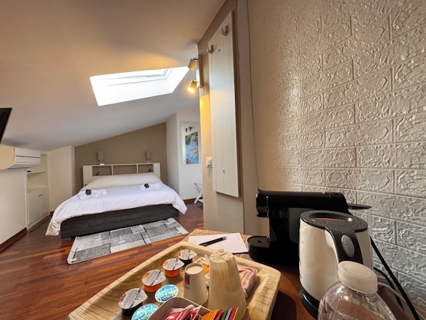 Quarto de hotel com uma cama, uma escrivaninha com cafeteira, um tabuleiro com alimentos e vista para o exterior através de um claraboia.