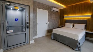 Quarto de hotel com uma cama, uma porta com o símbolo de acessibilidade para cadeiras de rodas, um aparelho de ar condicionado e uma mesinha de cabeceira.