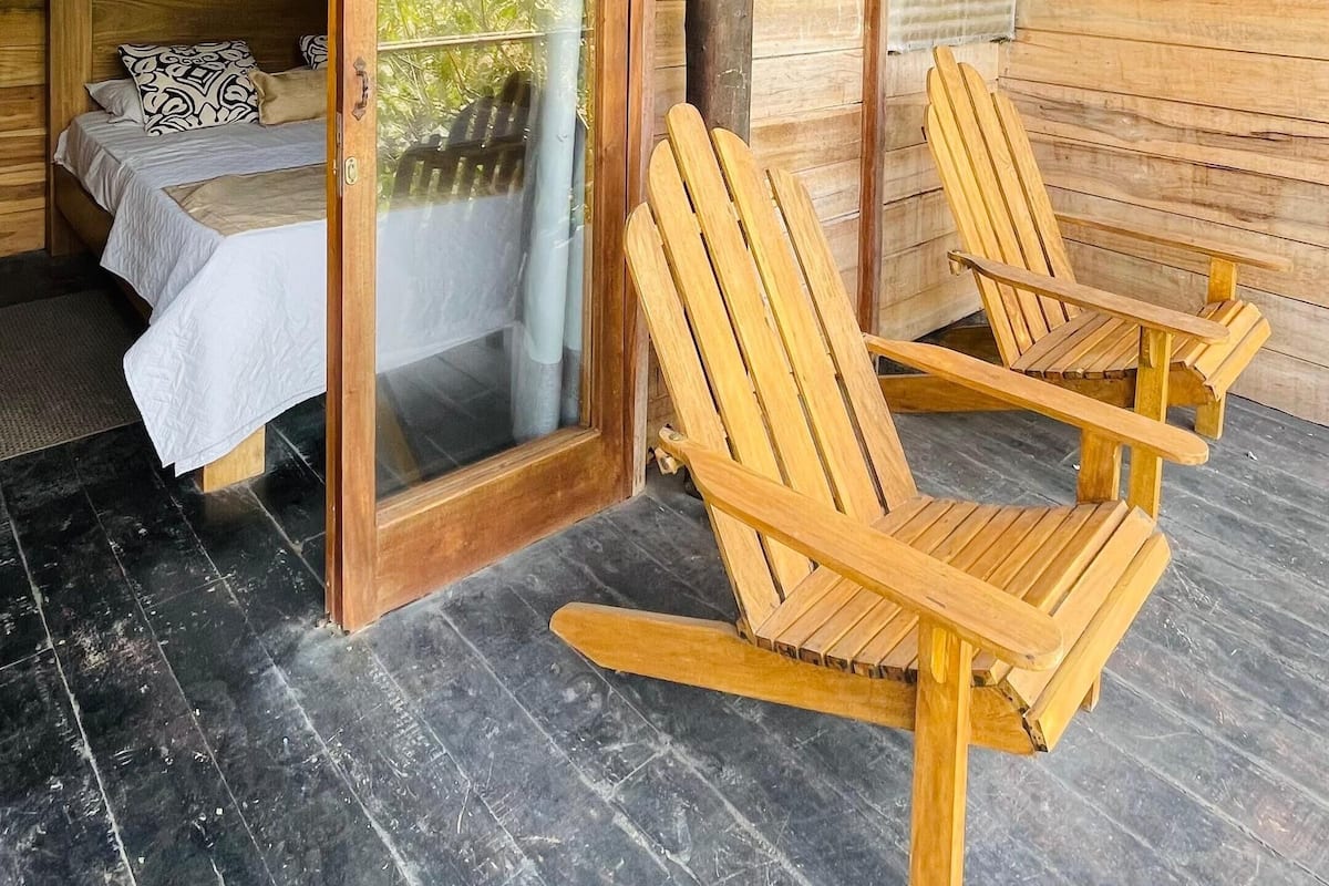 Uma cabana de madeira com telhado de palha, uma cama com cobertura branca e duas cadeiras de balanço de madeira sobre um piso de azulejos.