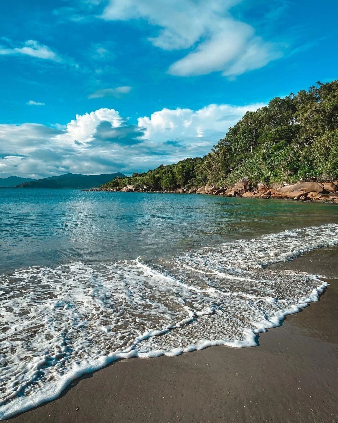 Uma praia com águas azuis cristalinas, ondas suaves beijando a costa, e um morro coberto por vegetação ao fundo.