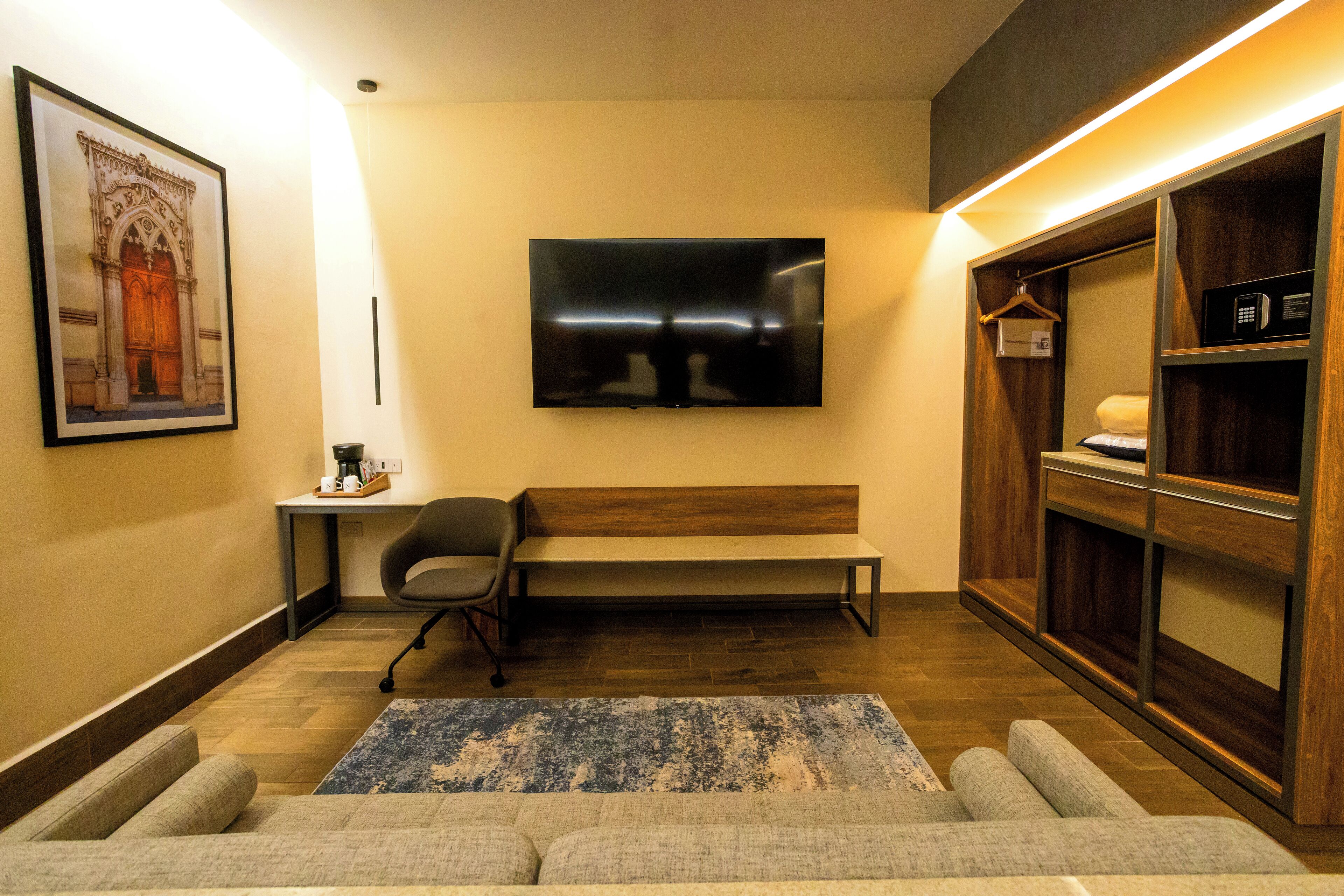 Quarto de hotel moderno com um banco de madeira, um sofá, uma TV fixada na parede, um cabideiro e um guarda-roupa de madeira.