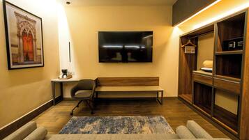 Quarto de hotel moderno com um banco de madeira, um sofá, uma TV fixada na parede, um cabideiro e um guarda-roupa de madeira.