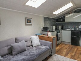 Um espaço de estar compacto com sofá cinzento, uma pequena mesa de madeira, área de cozinha com eletrodomésticos modernos e um claraboia acima do sofá.