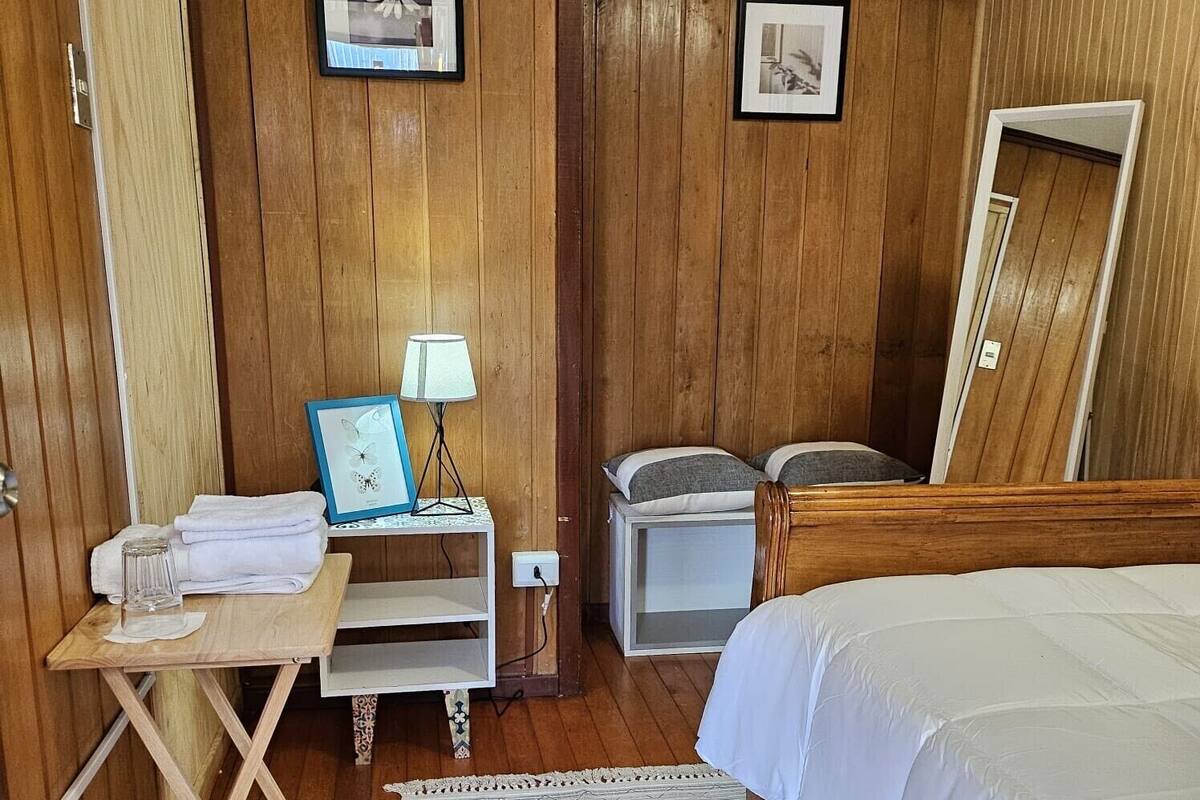 Um quarto com painéis de madeira, uma cama, uma mesinha de cabeceira com abajur, uma pequena mesa com toalhas e copos, e um espelho na parede.
