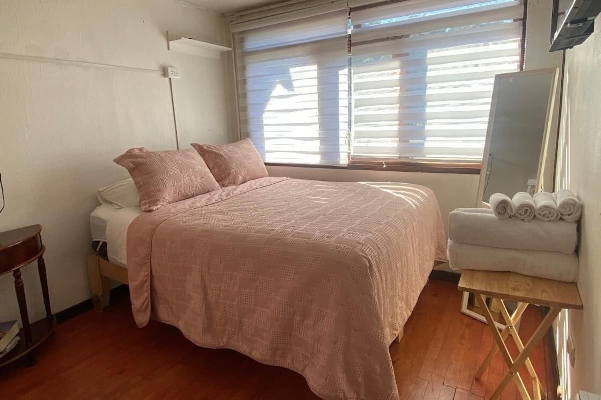 Quarto com cama, mesa de cabeceira, espelho e janela com persianas.