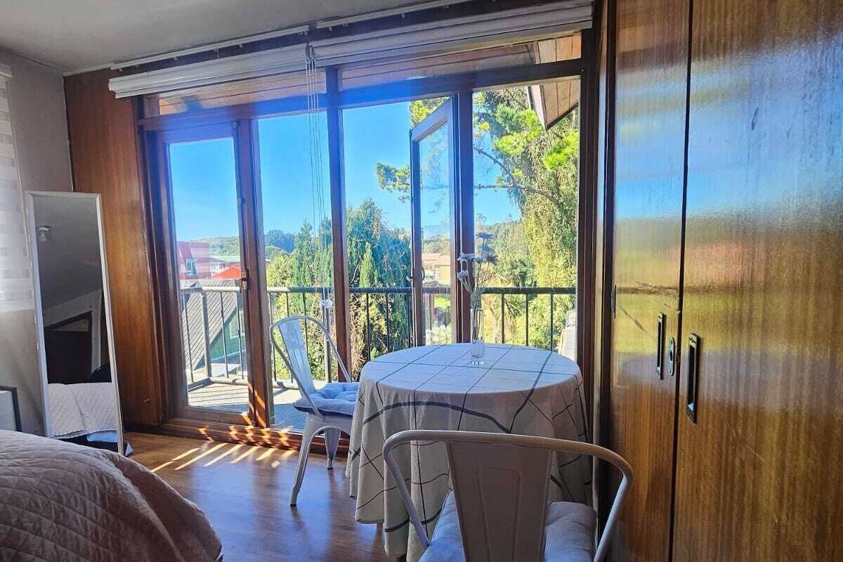 Quarto com cama grande, televisão, varanda com mesa e cadeiras, e vista para o exterior.