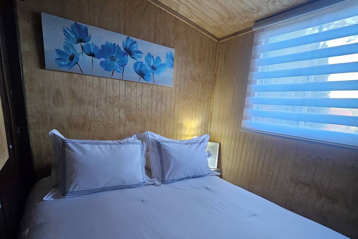 Um quarto com teto de madeira, cama com roupa de cama branca e um quadro de flores azuis na parede.