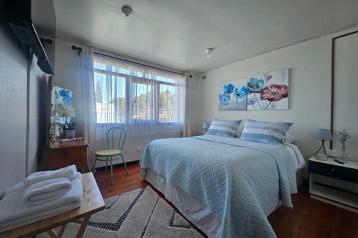 Quarto com cama, uma cadeira, mesa com toalhas, um quadro na parede e janela com cortinas.