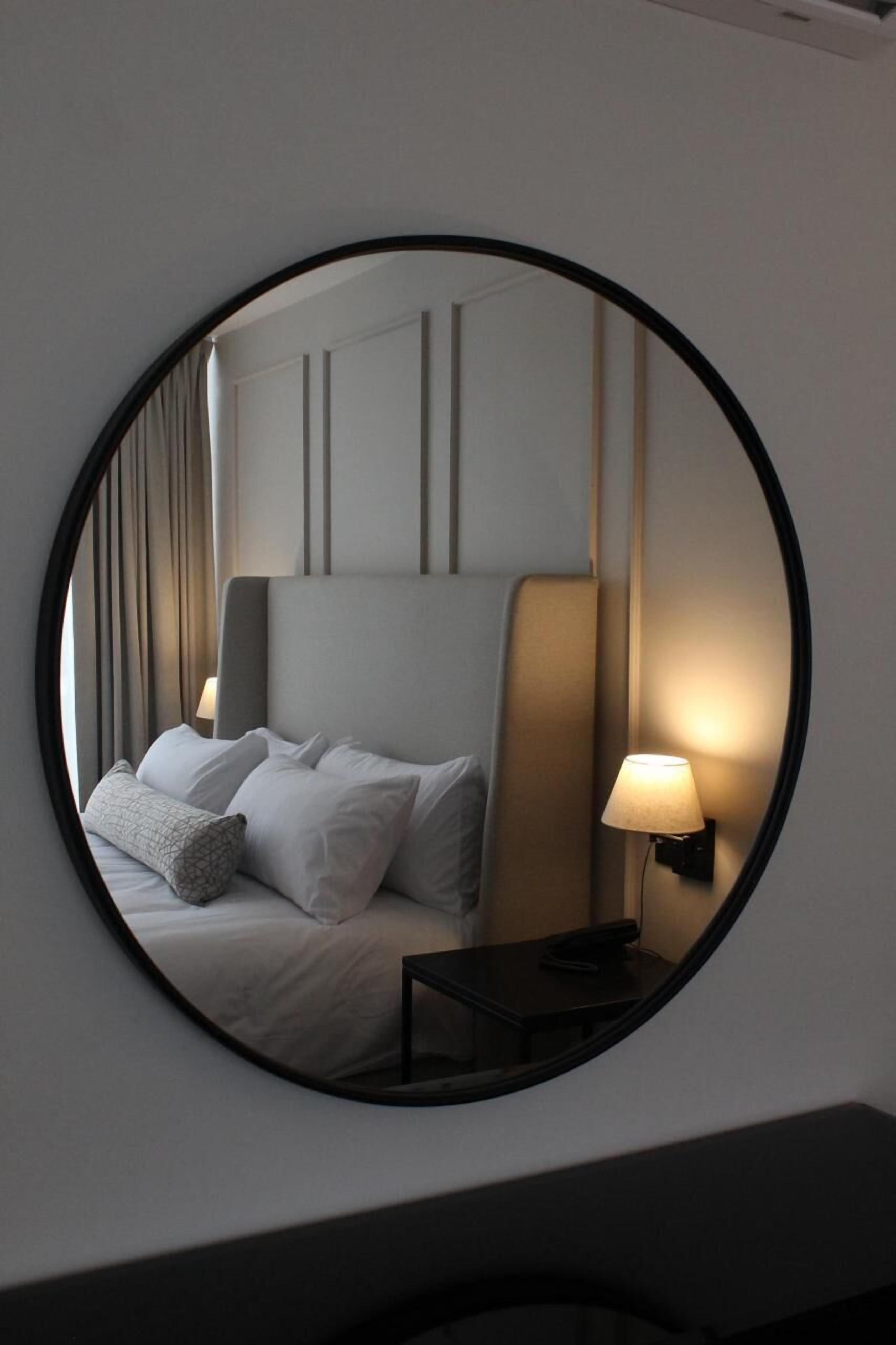 Um espelho circular reflete uma cama bem arrumada com almofadas e uma mesa de cabeceira com abajur.